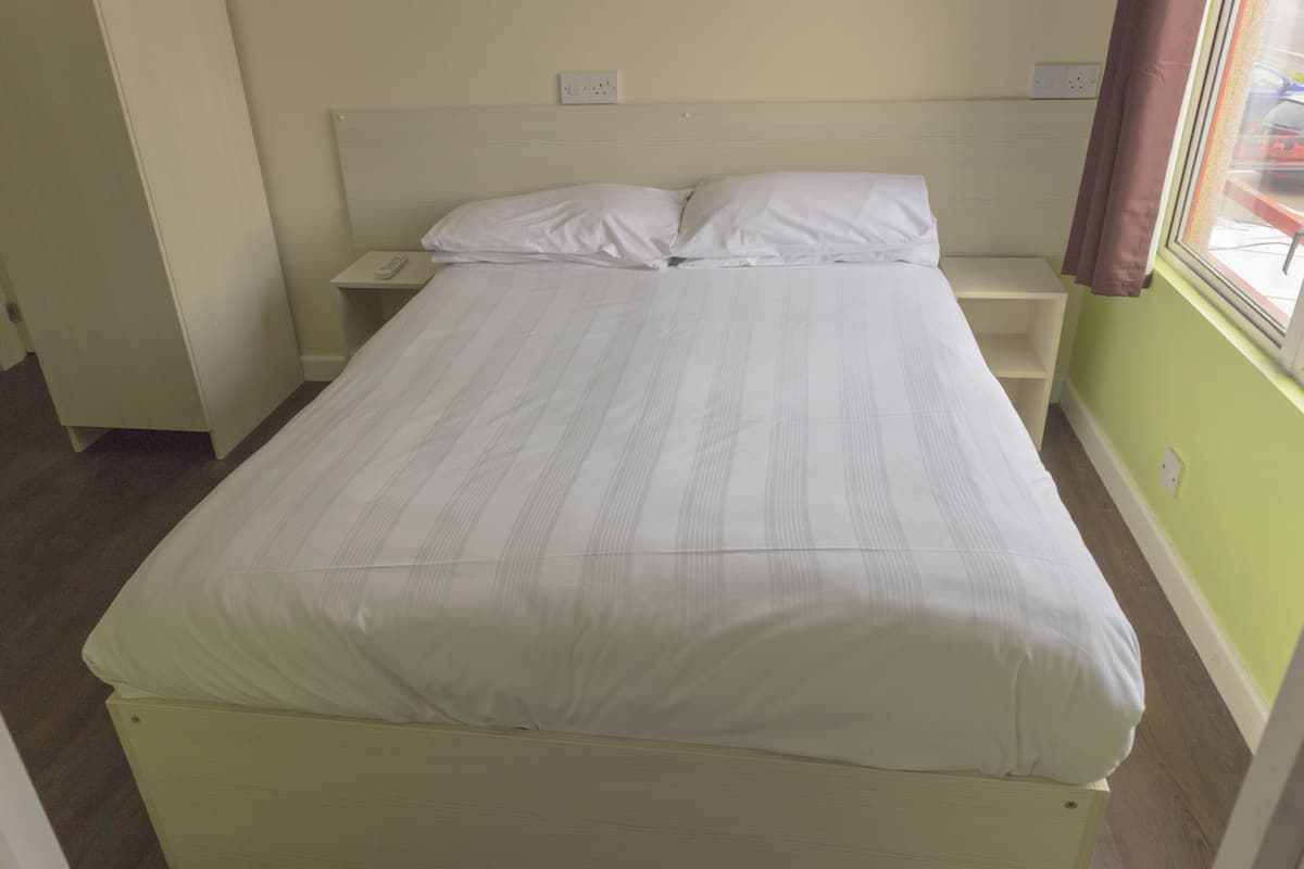 Cama com roupa de cama branca, mesa de cabeceira e janela com cortinas.