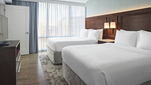 Quarto de hotel com duas camas, uma escrivaninha, uma luminária e vista da cidade por grandes janelas.