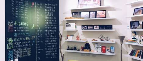 Um interior moderno com um quadro negro exibindo cronogramas de eventos, prateleiras com livros e objetos decorativos, e uma cadeira verde com um travesseiro com a inscrição 'Boa sorte'.