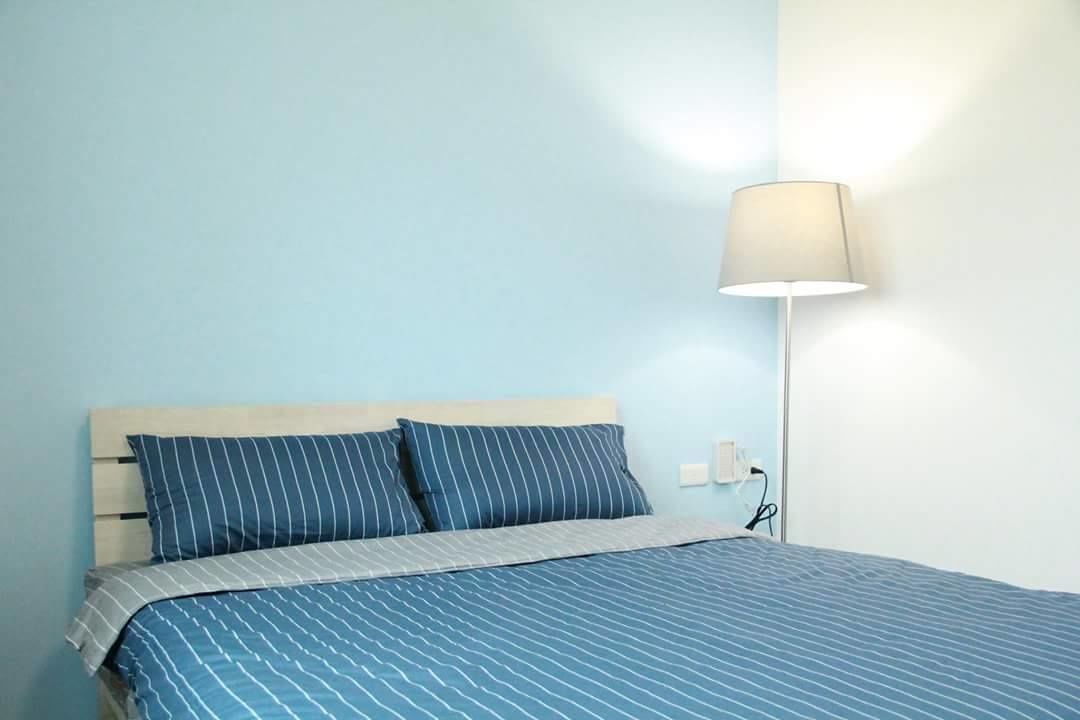 Cama com colcha listrada azul e almofadas, luminária branca e interruptor na parede.