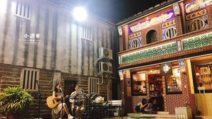 Um pátio externo com palco para música ao vivo, rodeado por edifícios com arquitetura tradicional e uma pequena área de jardim.
