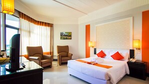 Quarto de hotel com uma cama grande, duas poltronas, uma escrivaninha com um computador e uma televisão.