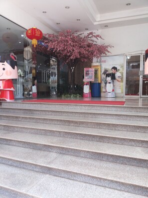 Entrada de um shopping com uma escadaria, uma grande árvore rosa e um lança-luzes vermelho.