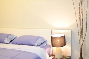 Cama arrumada com roupa de cama azul, luminária de cabeceira e um vaso com galhos em uma mesa de cabeceira.