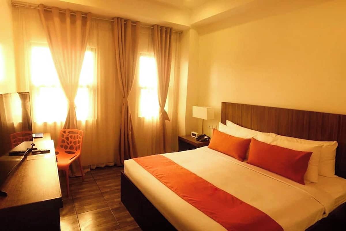 Quarto de hotel com uma cama grande, uma escrivaninha com cadeira, mesa de cabeceira com abajur e cortinas.