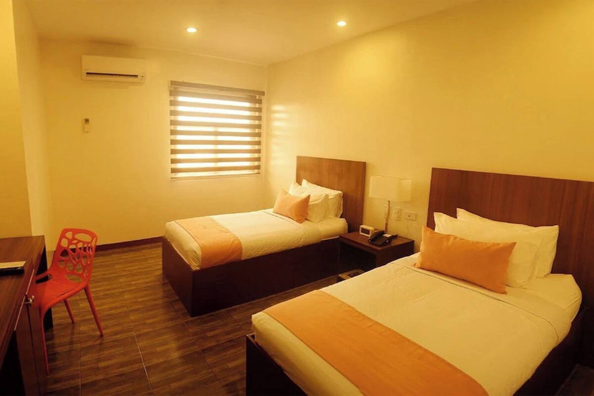 Quarto de hotel com duas camas, uma escrivaninha, uma cadeira e uma janela com persianas.