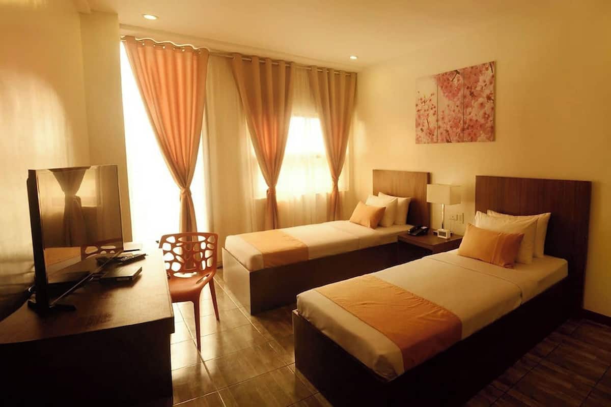 Quarto de hotel com duas camas de solteiro, uma escrivaninha com computador e cadeira, televisão e um quadro na parede.