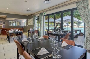 Restaurante com mesas preparadas para refeições, grandes janelas com vista para uma área de piscina.