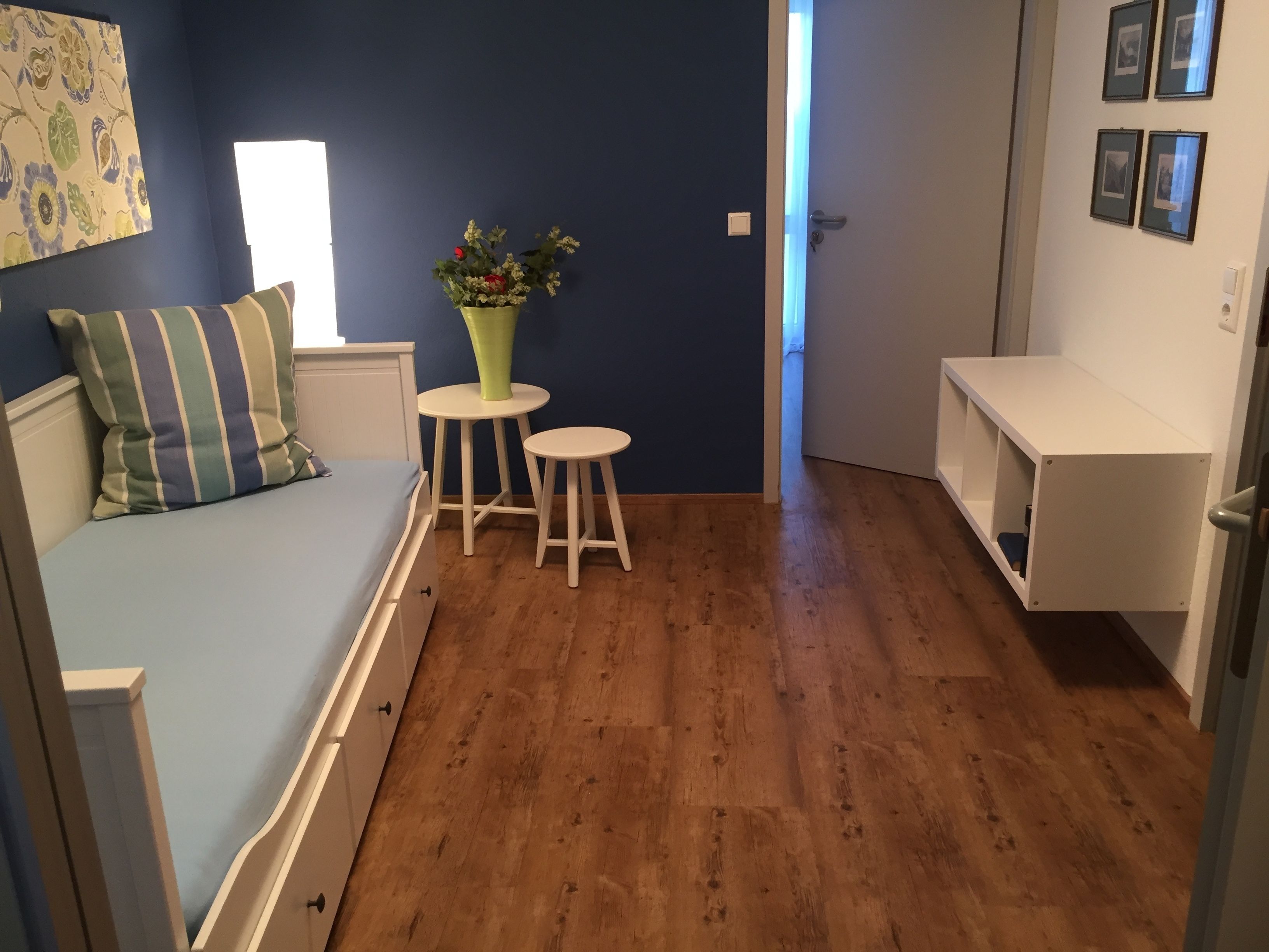 Um quarto pequeno com uma cama, uma mesinha com um vaso de flores e um armário branco.