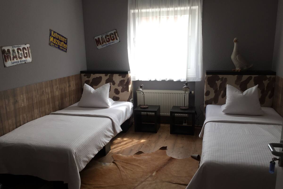 Quarto com duas camas, janela com cortinas leves, radiador e decorações nas paredes, incluindo placas para 'Kathreiners Malzkaffee' e 'MAGGI'.