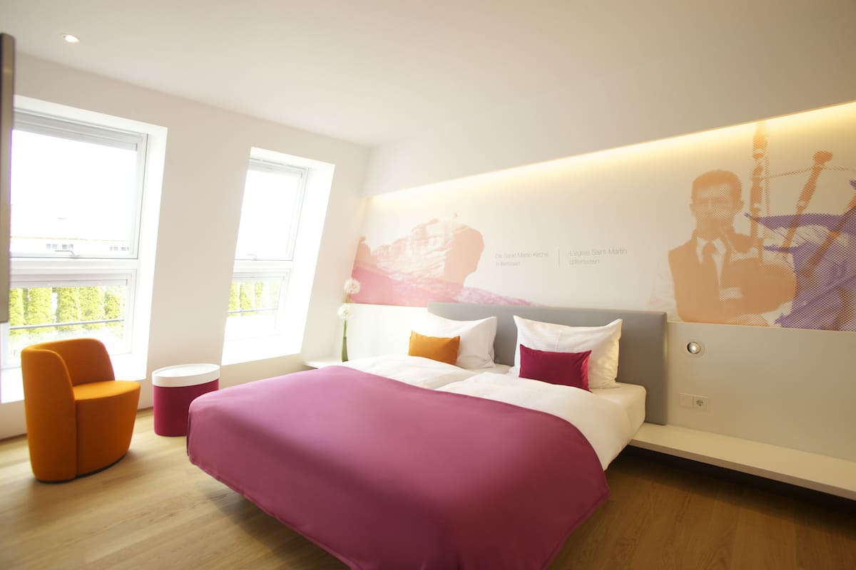 Quarto de hotel moderno com uma cama grande, área de estar com uma poltrona e janela com vista.