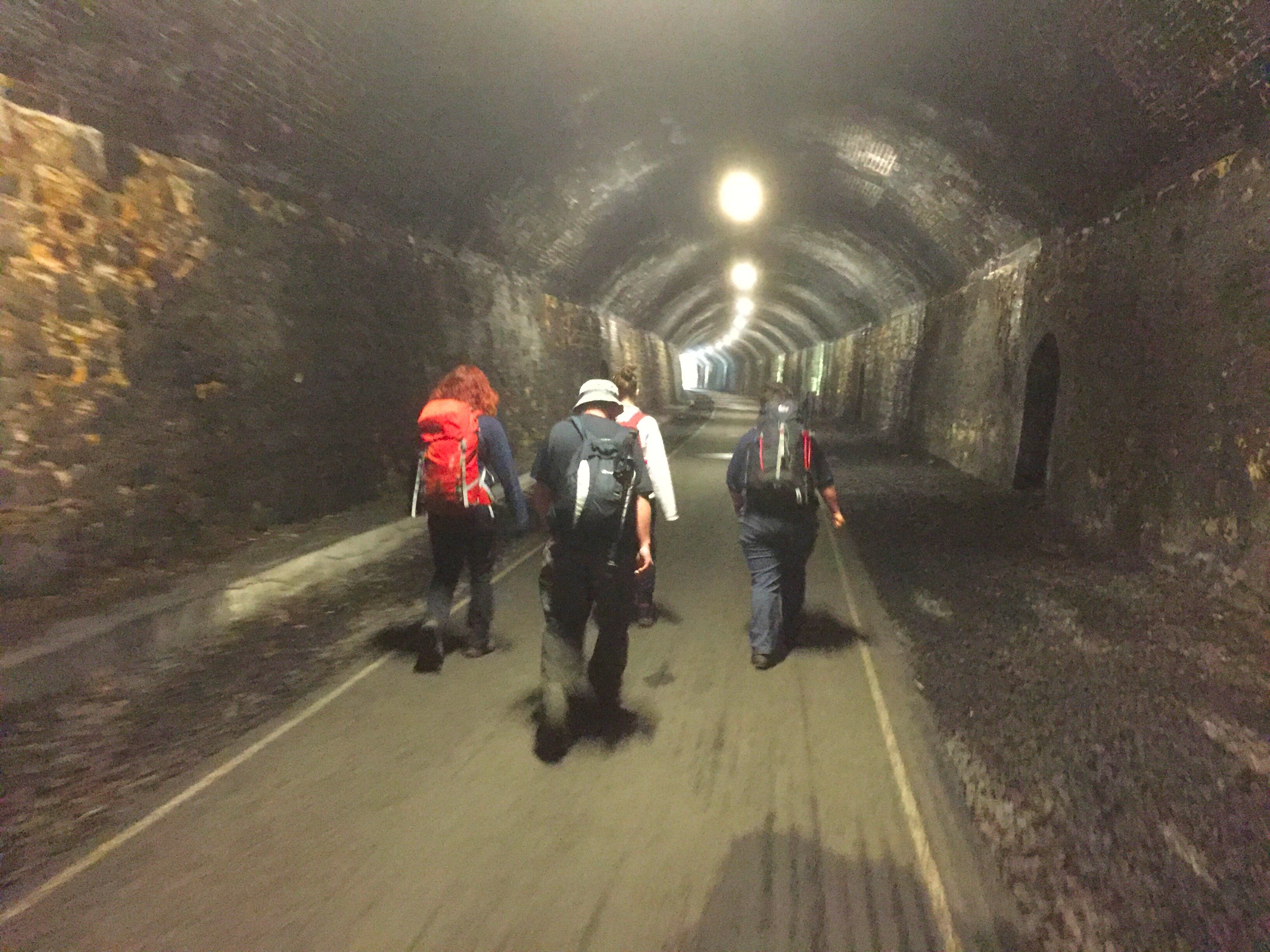 Um túnel com teto curvo e paredes, uma única fonte de luz no final, e três pessoas caminhando.