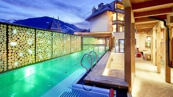 Uma piscina infinita com um deck de madeira, uma parede de pedra com padrões circulares e vista para as montanhas ao fundo.