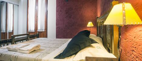 Quarto de hotel com uma cama, mesa de cabeceira, abajur, ventilador e janela com cortinas.