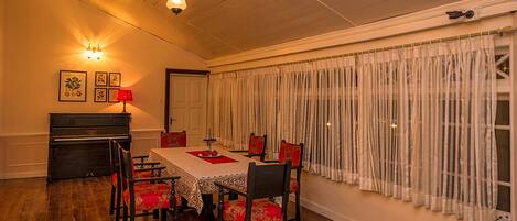 Uma sala de jantar com mesa de madeira, cadeiras estofadas vermelhas, um piano e uma janela com cortinas leves.