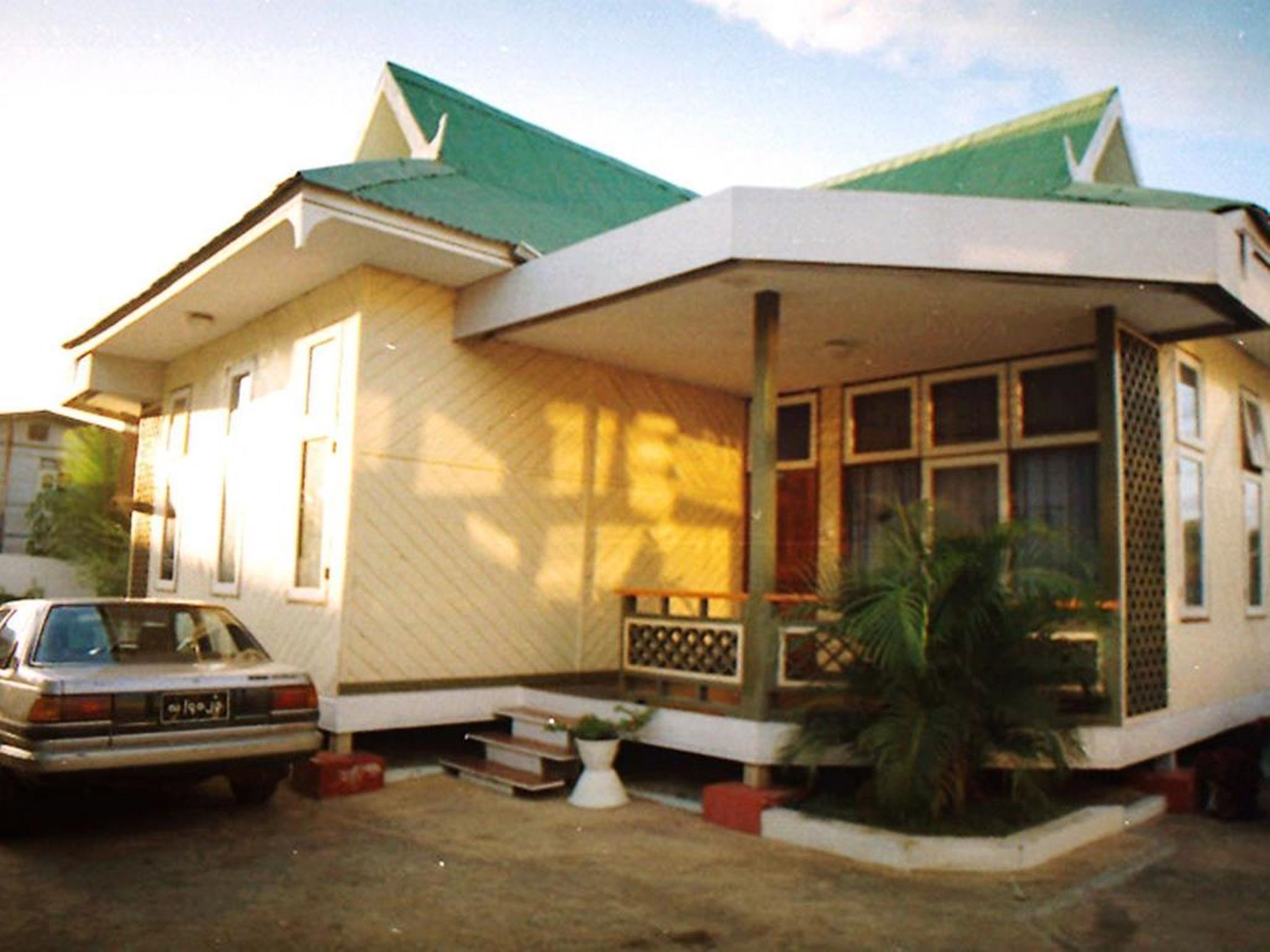 Uma casa de dois andares com telhado verde, varanda e um carro estacionado.