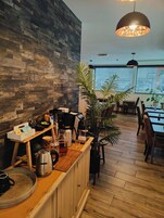 Um café acolhedor com balcão de madeira, máquinas de café e diversas bebidas. Há uma janela grande com persianas, um lustre pendente e um vaso de planta.