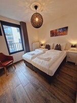 Quarto de hotel moderno com uma cama grande, uma cadeira vermelha, uma janela com vista para edifícios e um candeeiro no teto.
