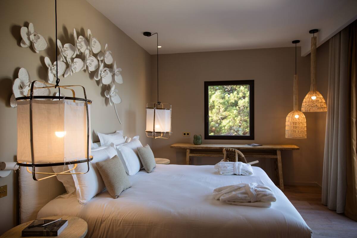 Quarto de hotel com uma cama grande, uma mesa de cabeceira, uma janela com vista para árvores e duas luzes pendentes.