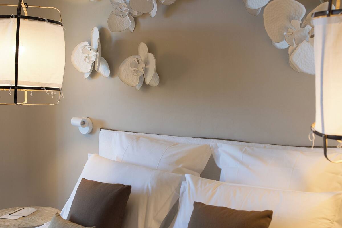 Quarto de hotel com cama grande, várias almofadas, decoração com orquídea na parede e luminária suspensa.