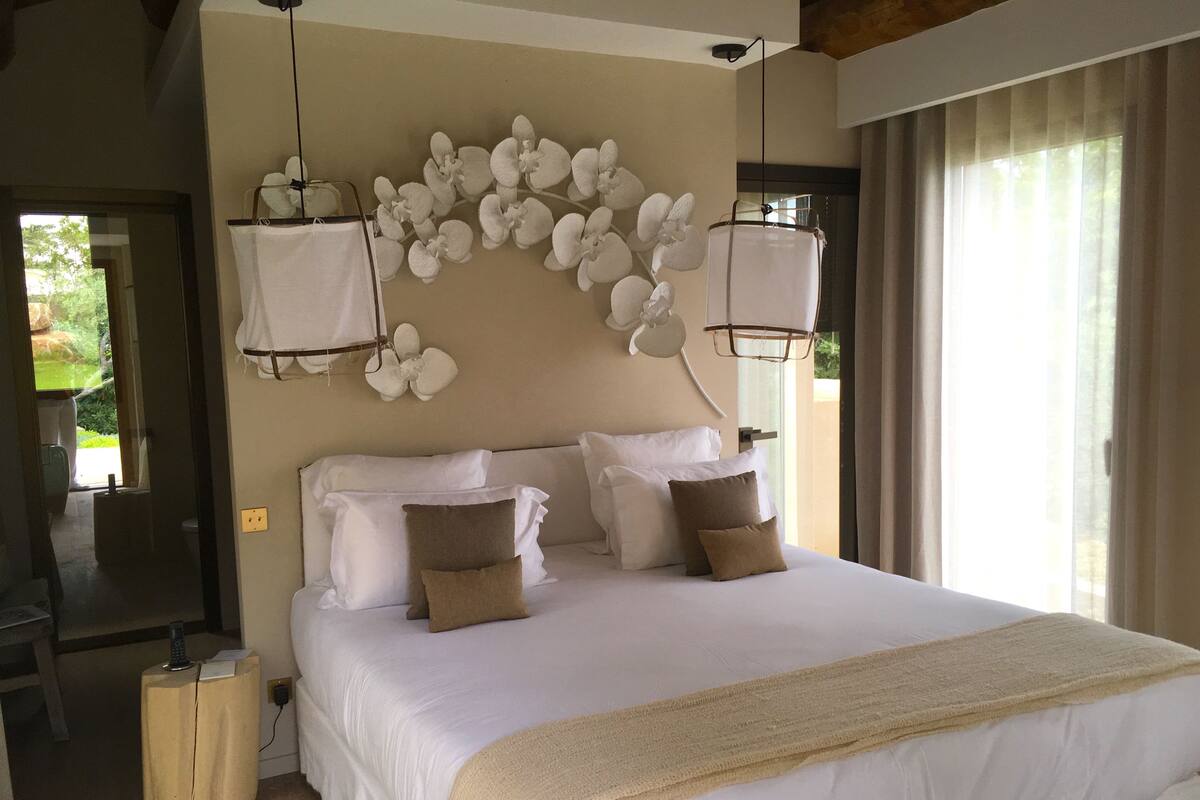 Uma cama grande com lençóis brancos e várias almofadas, uma mesa de cabeceira de madeira com um controle remoto e um candeeiro fixado na parede.