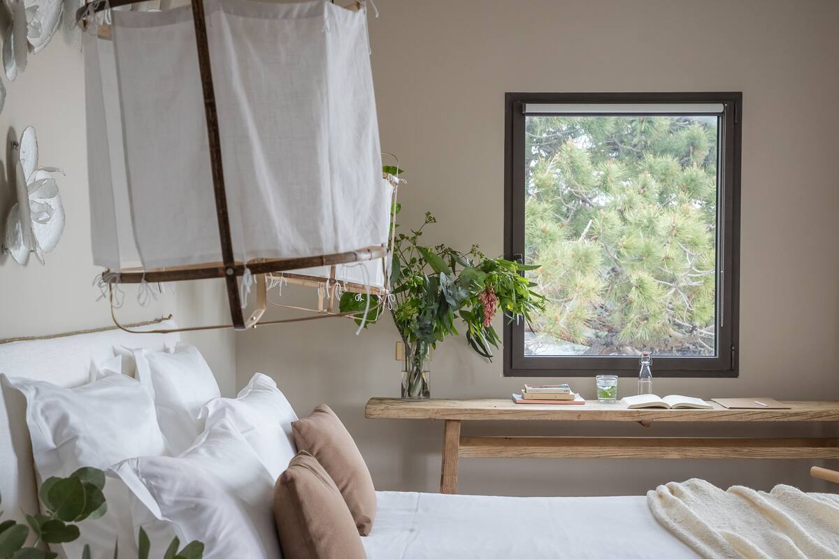 Quarto com cama, mesa de cabeceira e janela com vista para árvores.