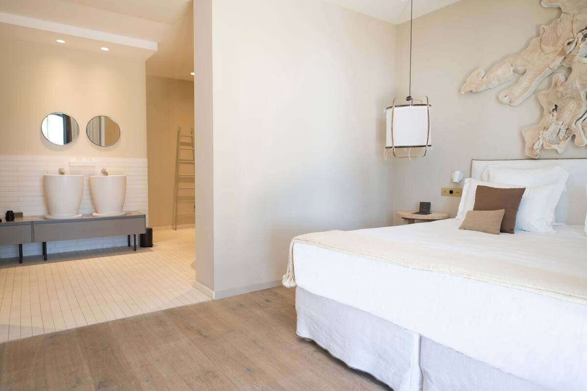 Quarto de hotel moderno com uma cama grande, duas mesas de cabeceira, um criado-mudo, um espelho e uma luminária fixada na parede.