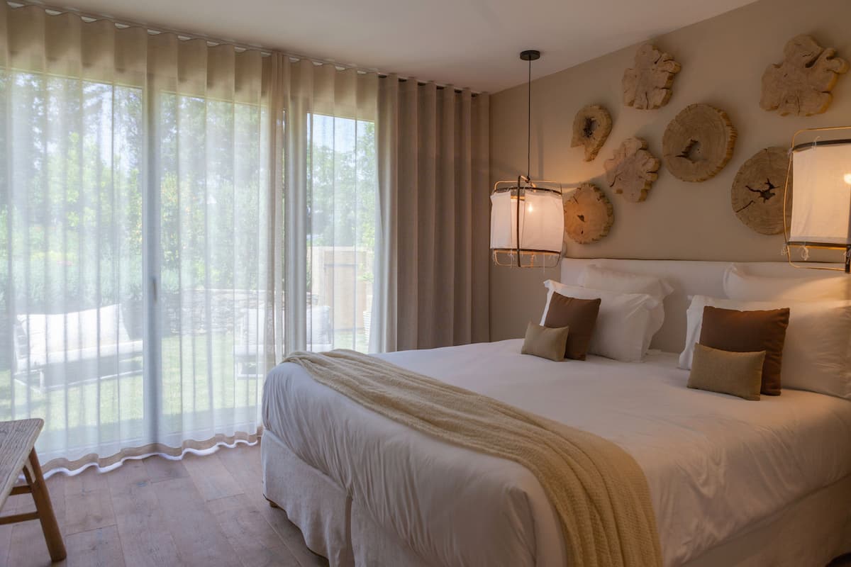 Uma cama grande com um cobertor bege, uma cadeira de madeira e vista para o verde através de cortinas leves.