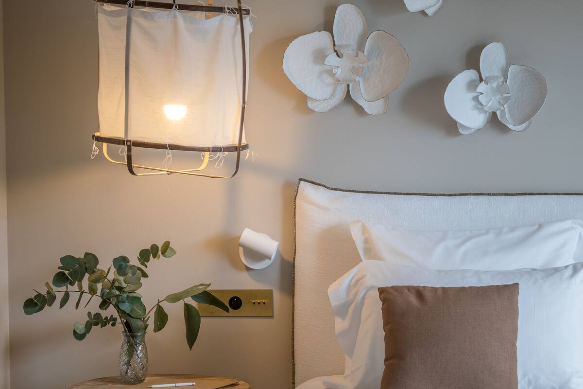 Quarto com cama, mesa de cabeceira com um vaso de plantas verdes, luminária fixada na parede e arte de parede com orquídeas brancas.