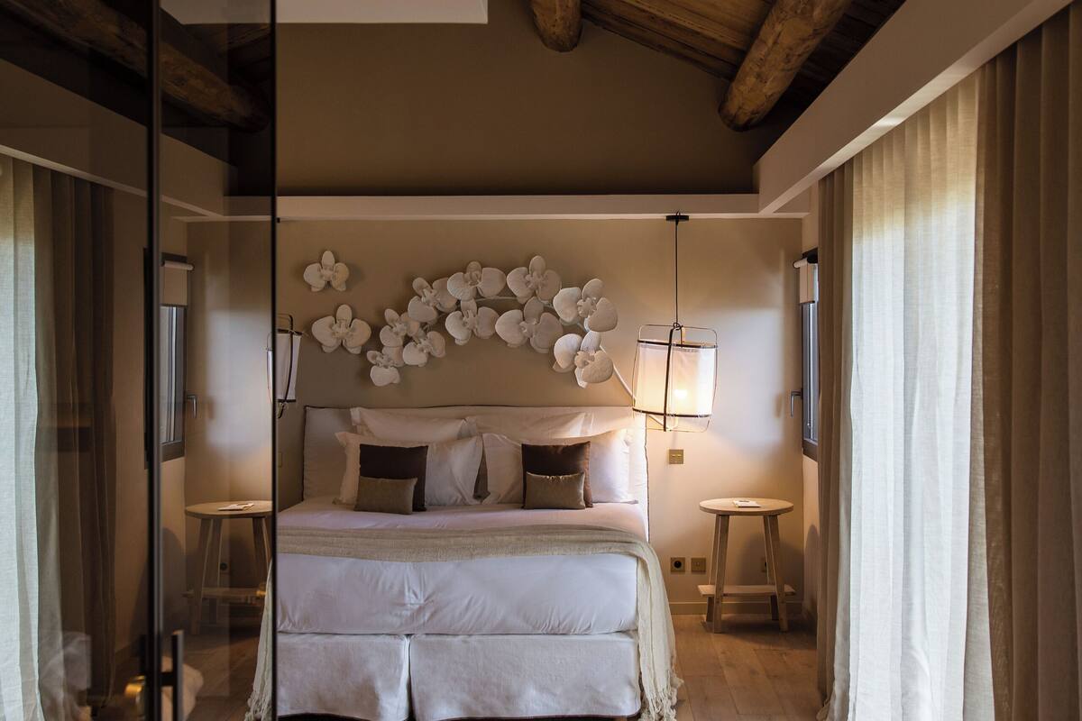 Quarto com teto de madeira, uma cama grande, luminária fixada na parede e divisória de vidro.