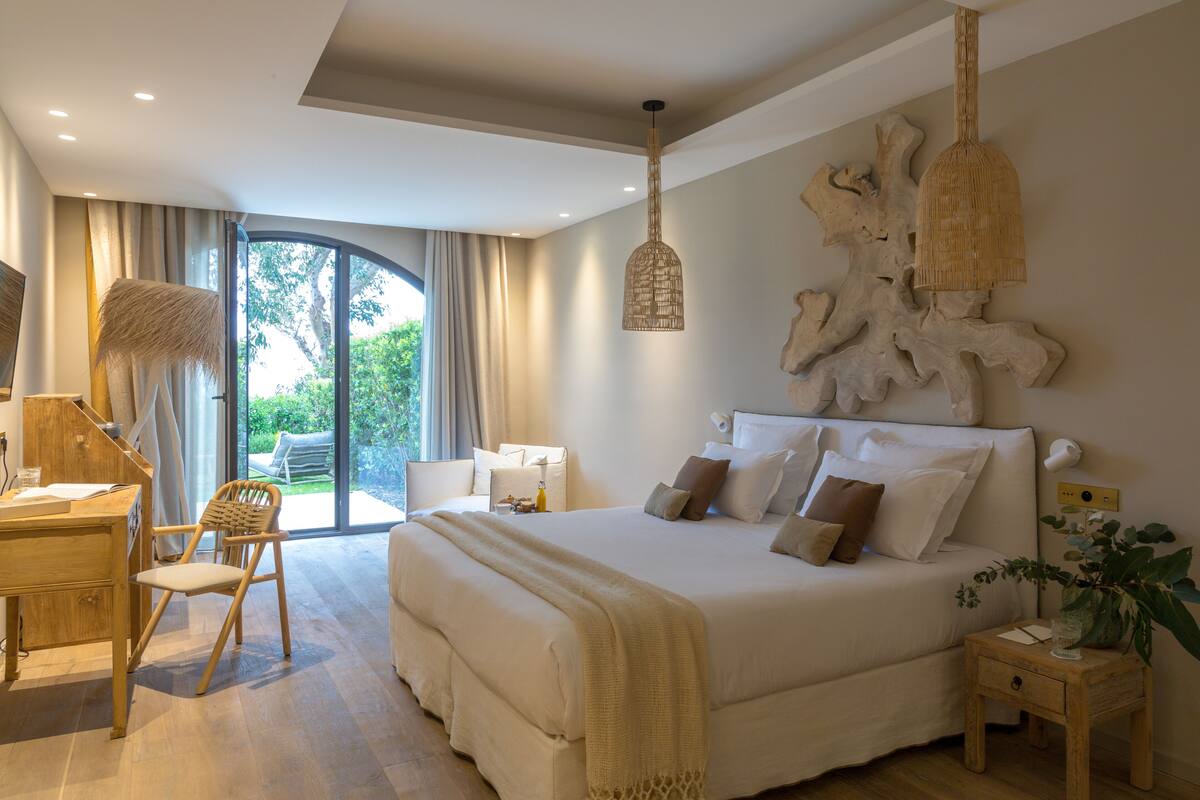 Quarto com uma cama grande, uma escrivaninha e uma cadeira. Há uma janela com cortinas, um lustre e uma planta suspensa na parede.