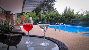 Uma mesa de vidro com duas bebidas e um vaso de planta. Ao fundo, uma piscina rodeada por árvores.