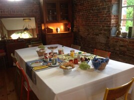 Uma mesa de jantar posta com diversos pratos e bebidas, rodeada por cadeiras em um cômodo com paredes de tijolo e uma janela.