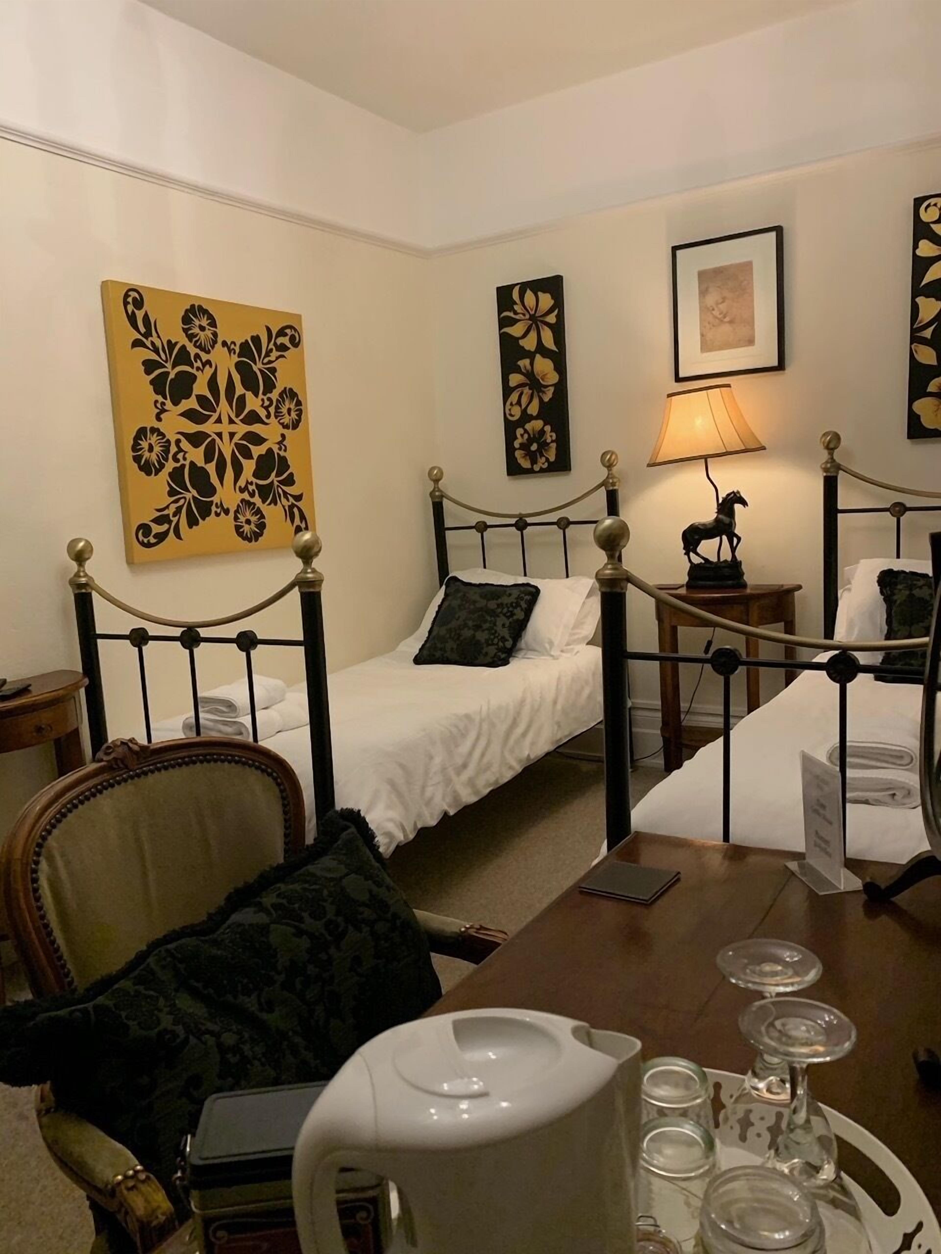 Quarto de hotel com duas camas de solteiro, uma mesa de madeira com jarra e copos brancos, uma cadeira e obras de arte nas paredes.