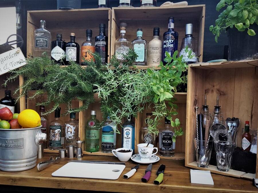 Uma área de bar com diversas garrafas de gin e outras bebidas, um balde de frutas e uma xícara de café.