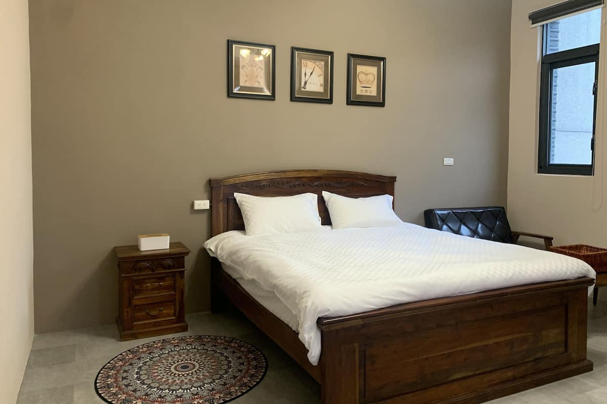 Quarto com cama de madeira, mesa de cabeceira, cadeira, tapete redondo estampado e três quadros na parede.