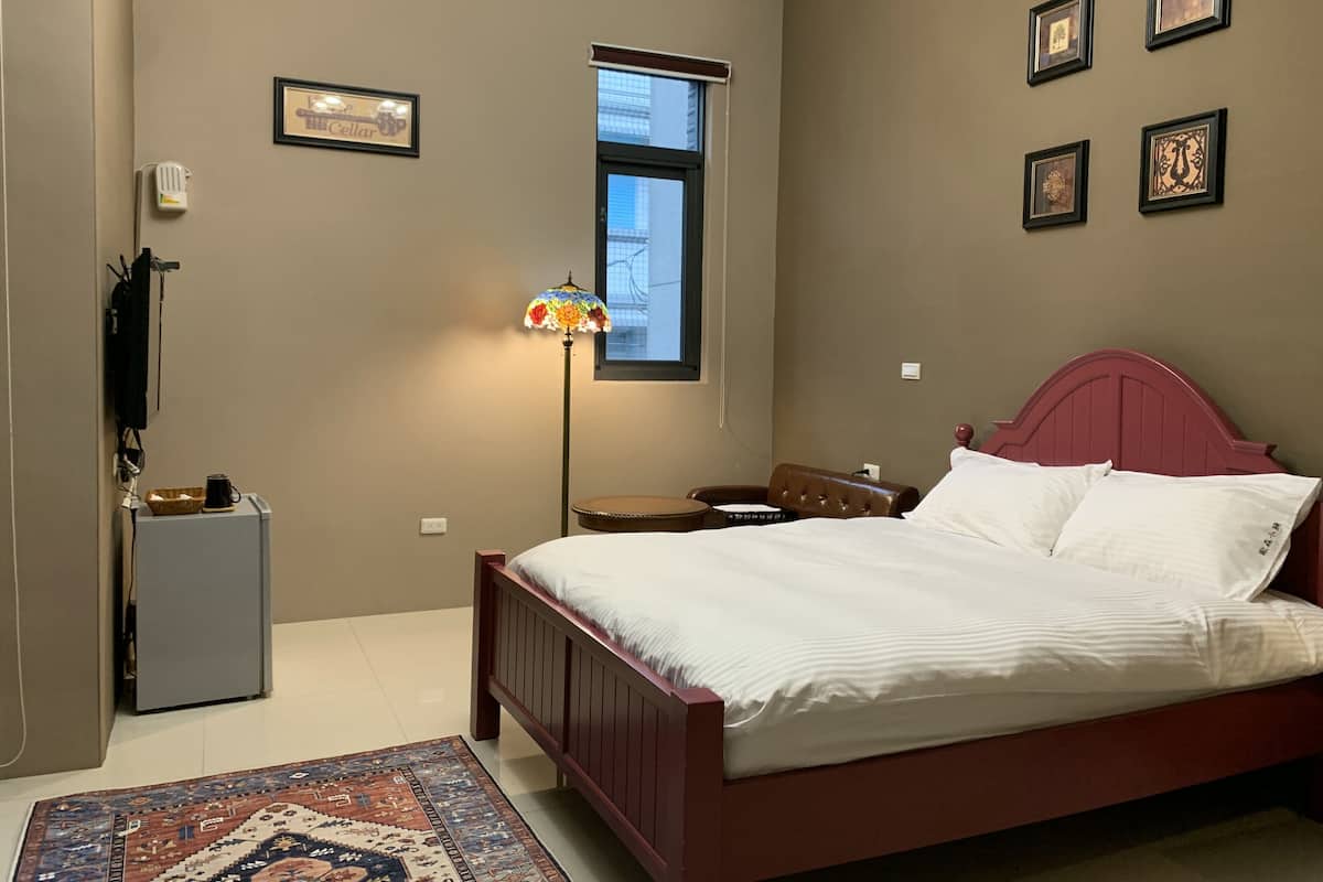 Quarto com cama, mesa de cabeceira, luminária, janela e televisão fixada na parede.