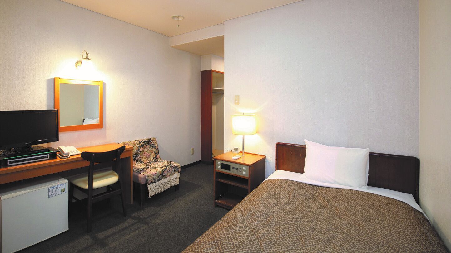 Quarto de hotel com cama, escrivaninha com televisão, cadeira, luminária de mesa e um pequeno frigorífico.
