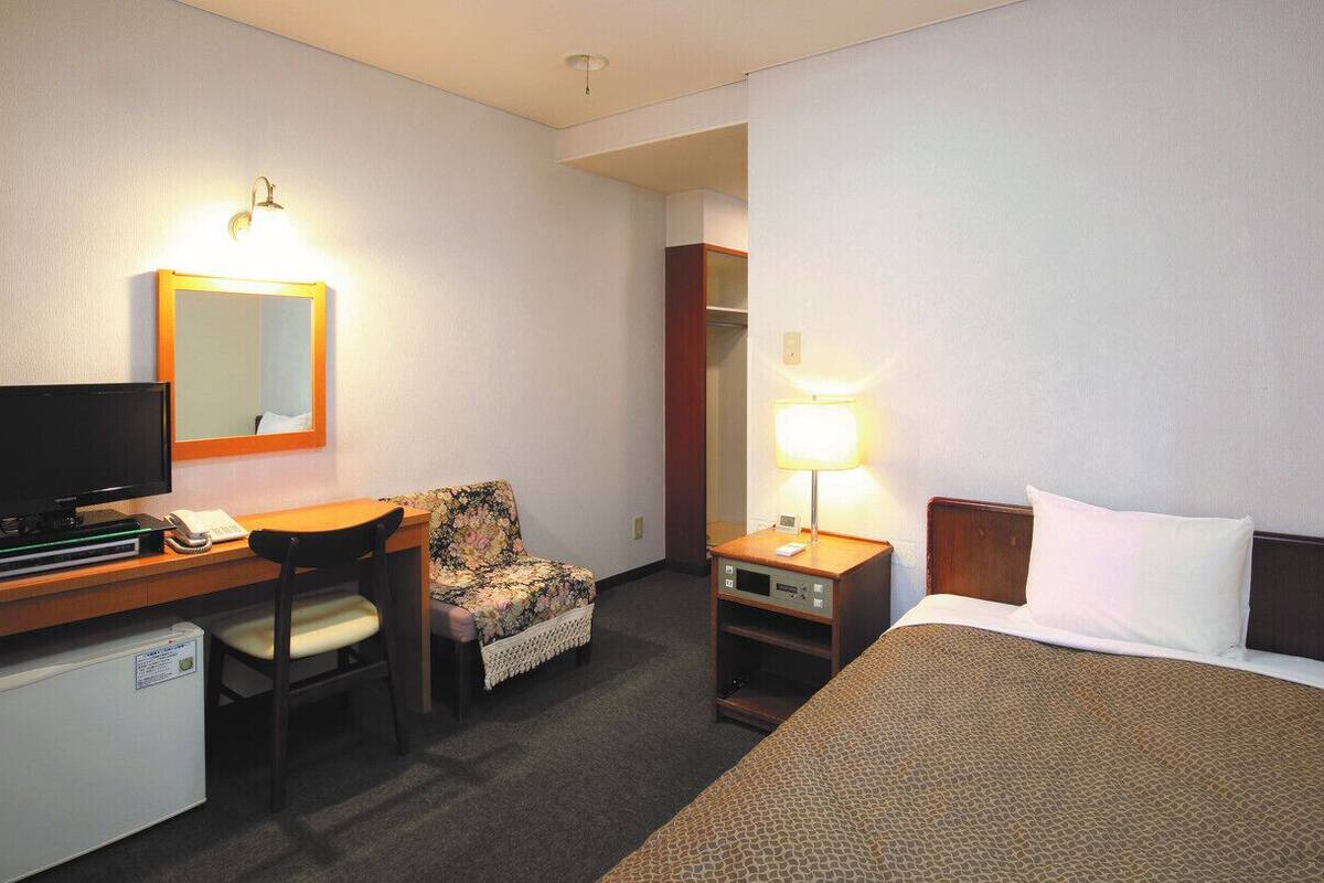 Quarto de hotel com cama, escrivaninha com televisão, cadeira, luminária de mesa e um pequeno frigorífico.