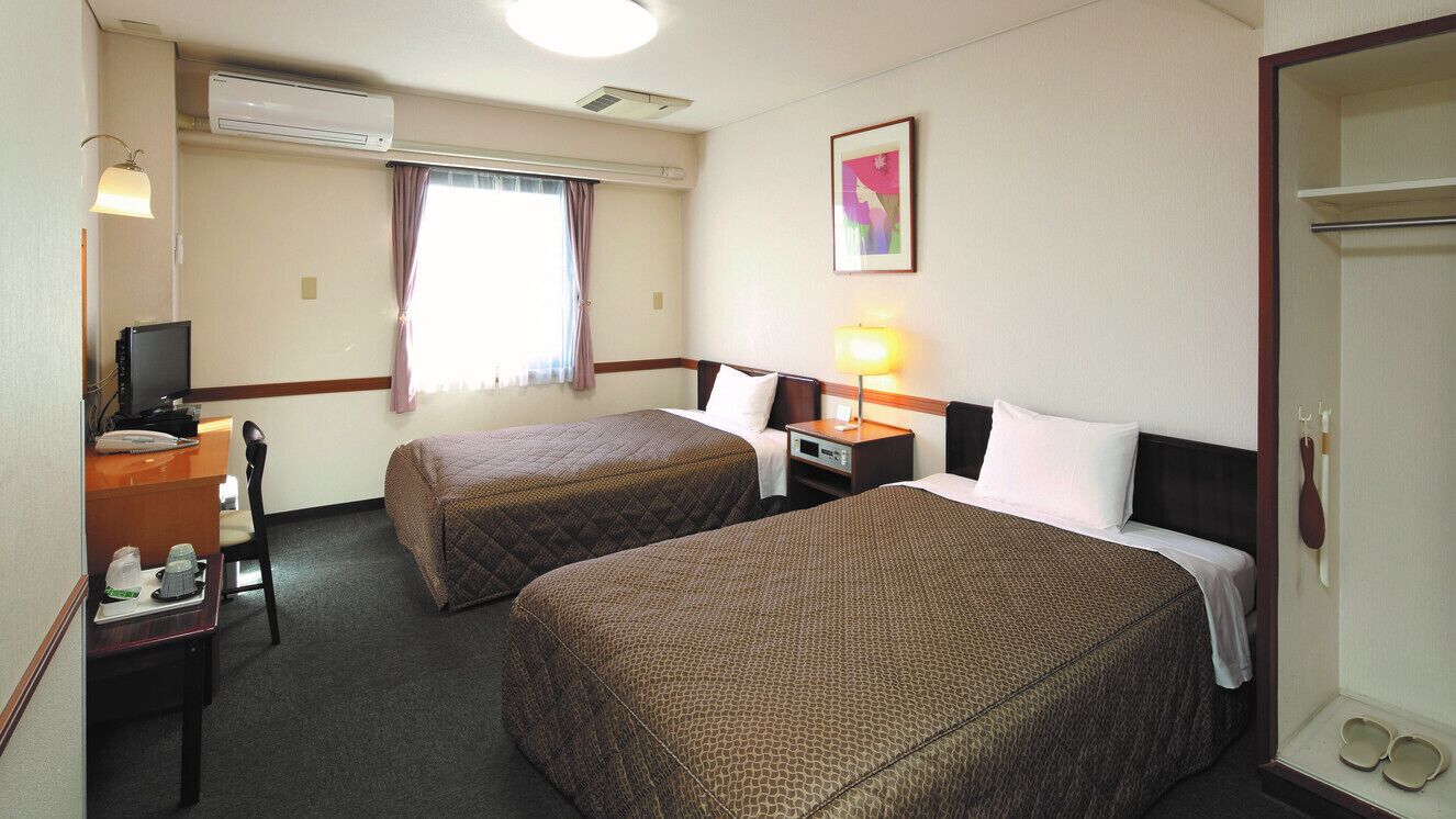 Quarto de hotel com duas camas, uma mesa com computador, uma cadeira, abajur e janela com cortinas.