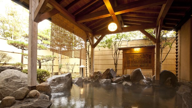 Uma onsen japonesa tradicional, com estrutura de madeira, banheira de pedra rodeada por pedras e cerca de bambu.
