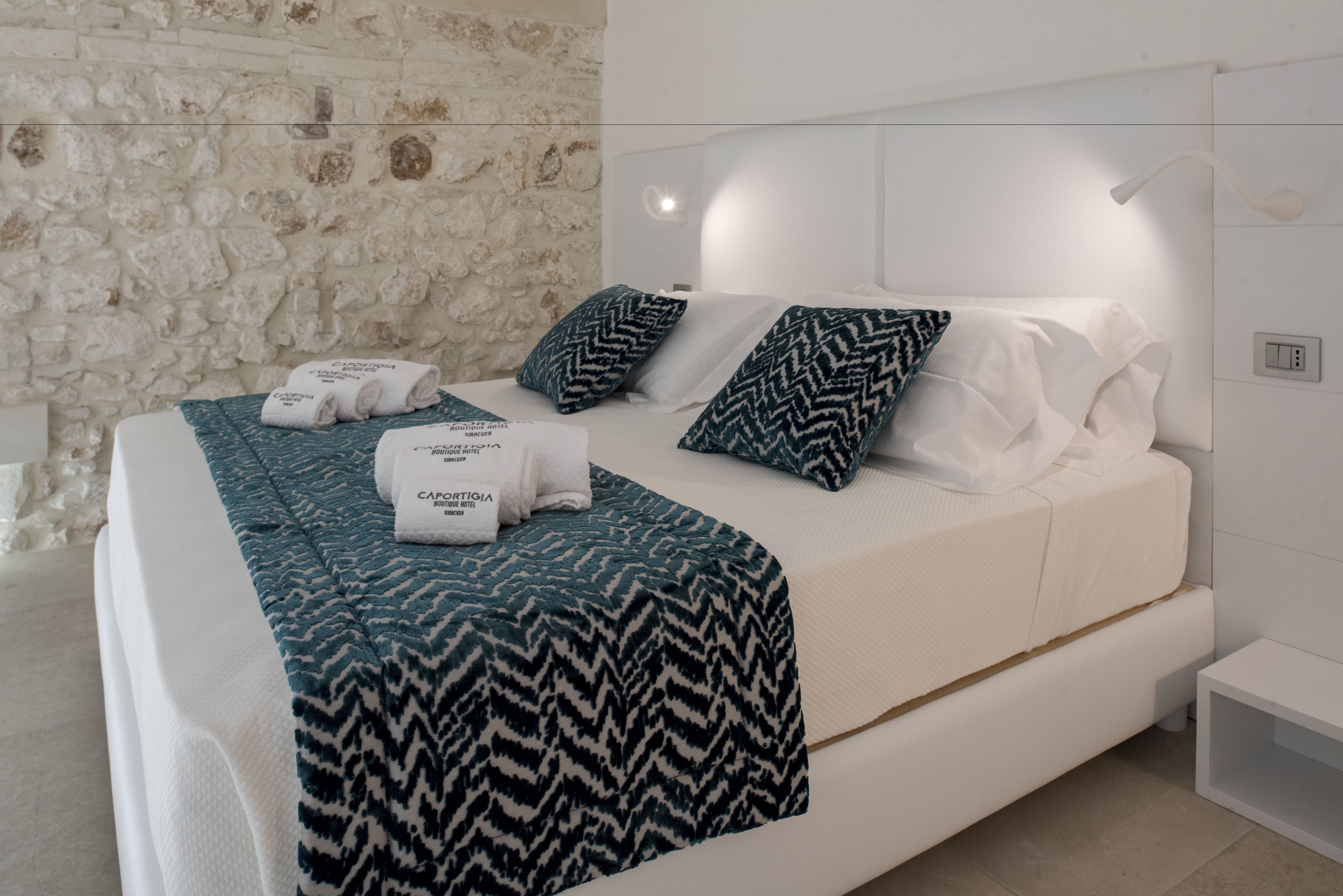 Quarto moderno com parede de pedra, cama branca e manta estampada em tons de verde-água e branco.