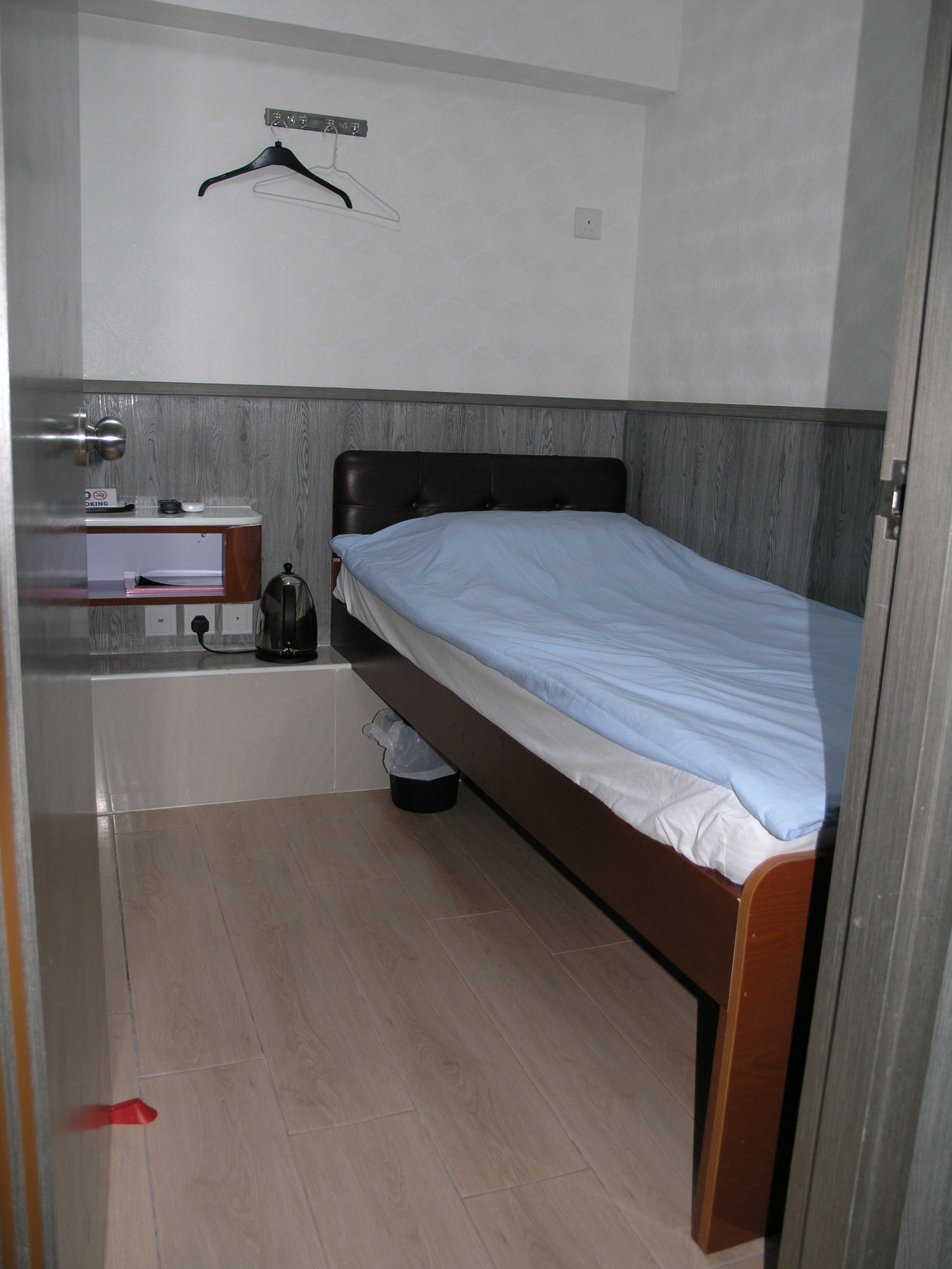 Quarto pequeno com uma cama de solteiro, cabeceira de madeira, mesa de cabeceira e um cabideiro.