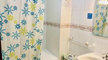 Casa de banho com cortina de chuveiro estampada com flores azuis e amarelas, pia branca com espelho em cima e prateleira verde com artigos de higiene pessoal.