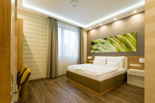 Quarto de hotel com uma cama grande, piso de madeira, janela com cortinas, um quadro pendurado na parede e uma mesa de cabeceira com abajur.