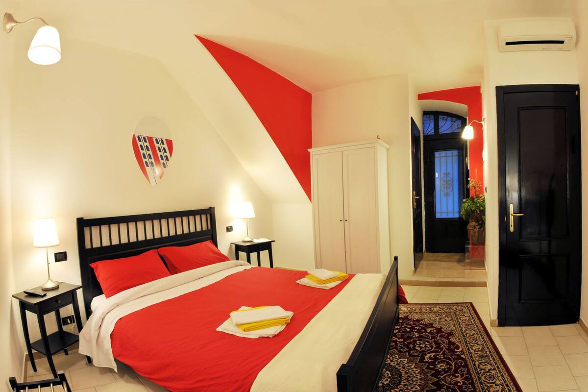 Quarto de hotel com esquema de cores vermelho e branco, cama com roupa de cama vermelha, cabeceira preta, duas mesas de cabeceira com luminárias, guarda-roupa branco e tapete estampado.