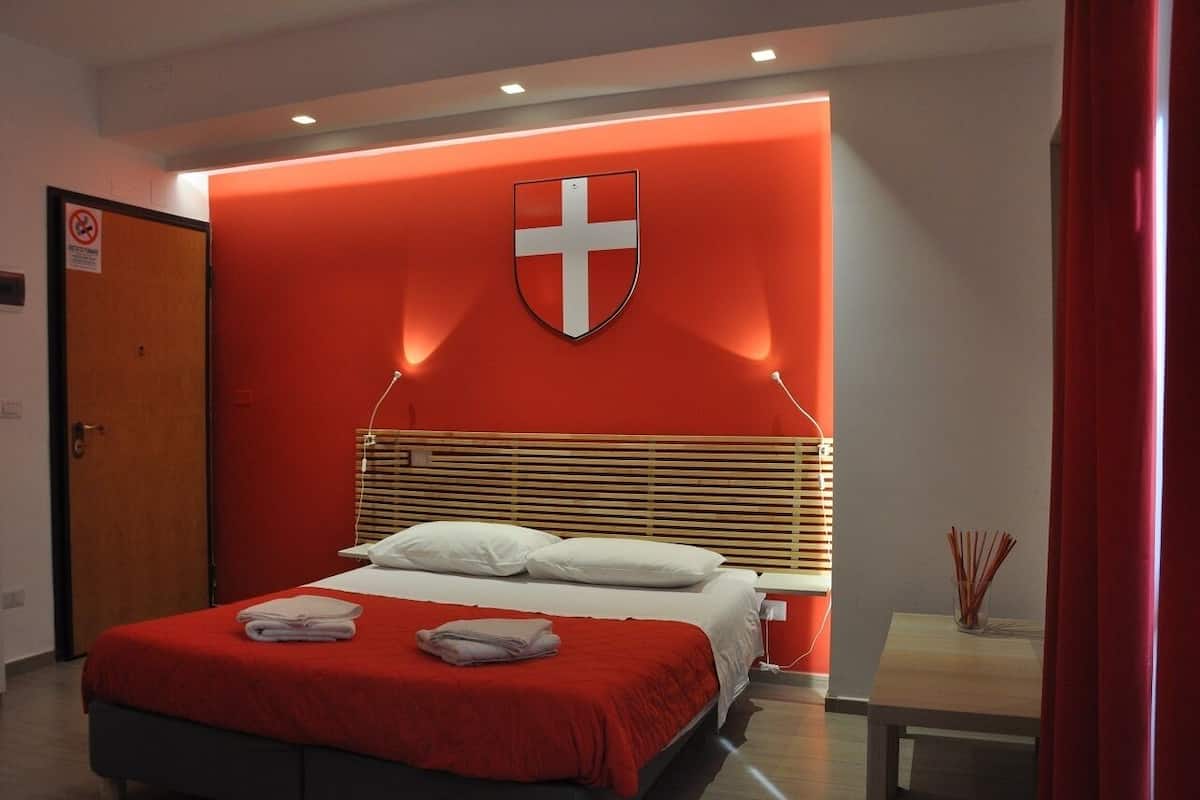Quarto de hotel com cama, cabeceira de madeira, parede vermelha e um brasão.