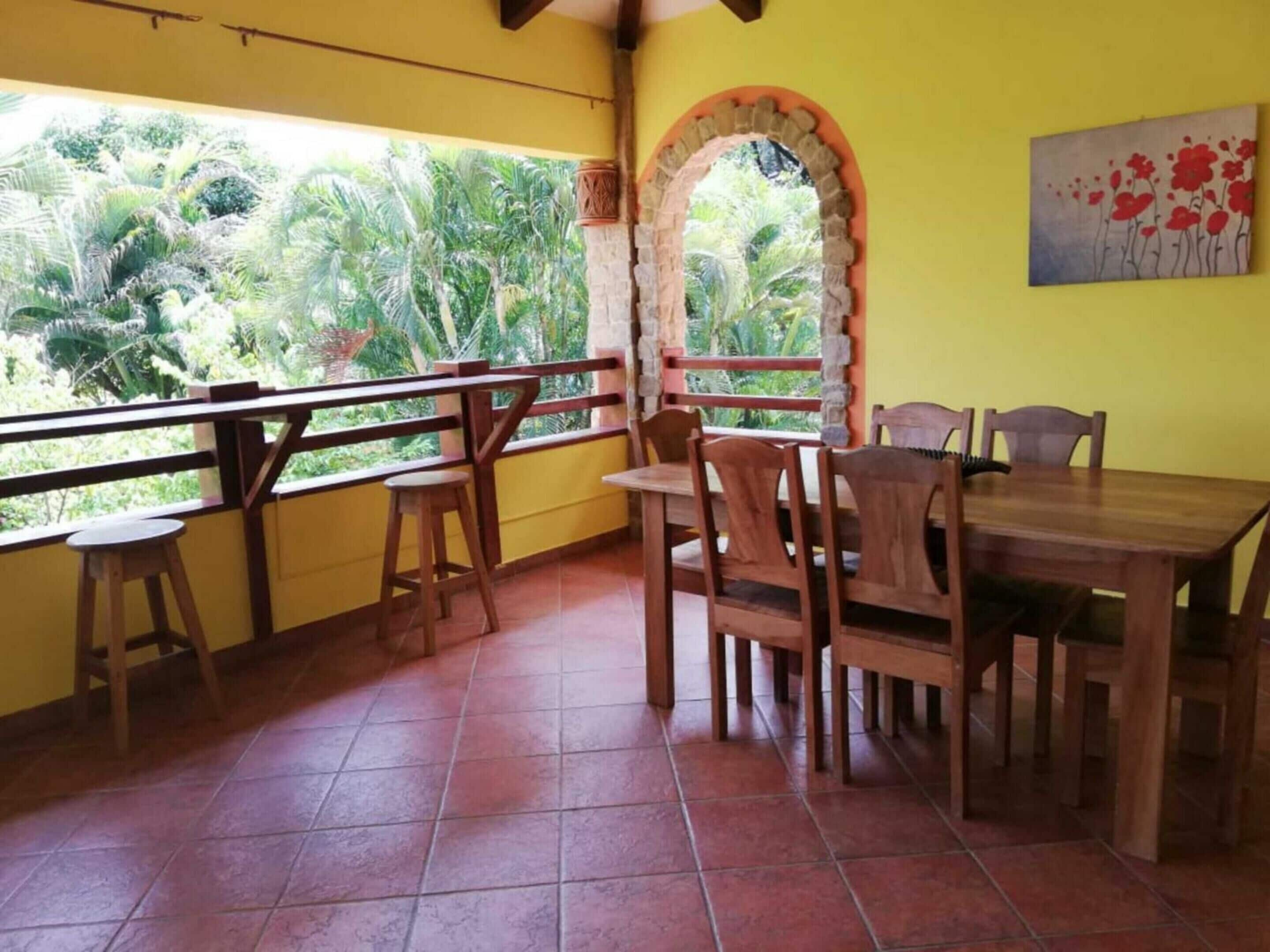 Uma sala de jantar com mesa e cadeiras de madeira, uma varanda com guarda-corpo e vista para o verde lá fora.