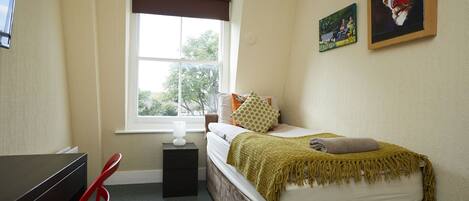 Quarto com cama, escrivaninha, cadeira, janela e duas pinturas na parede.