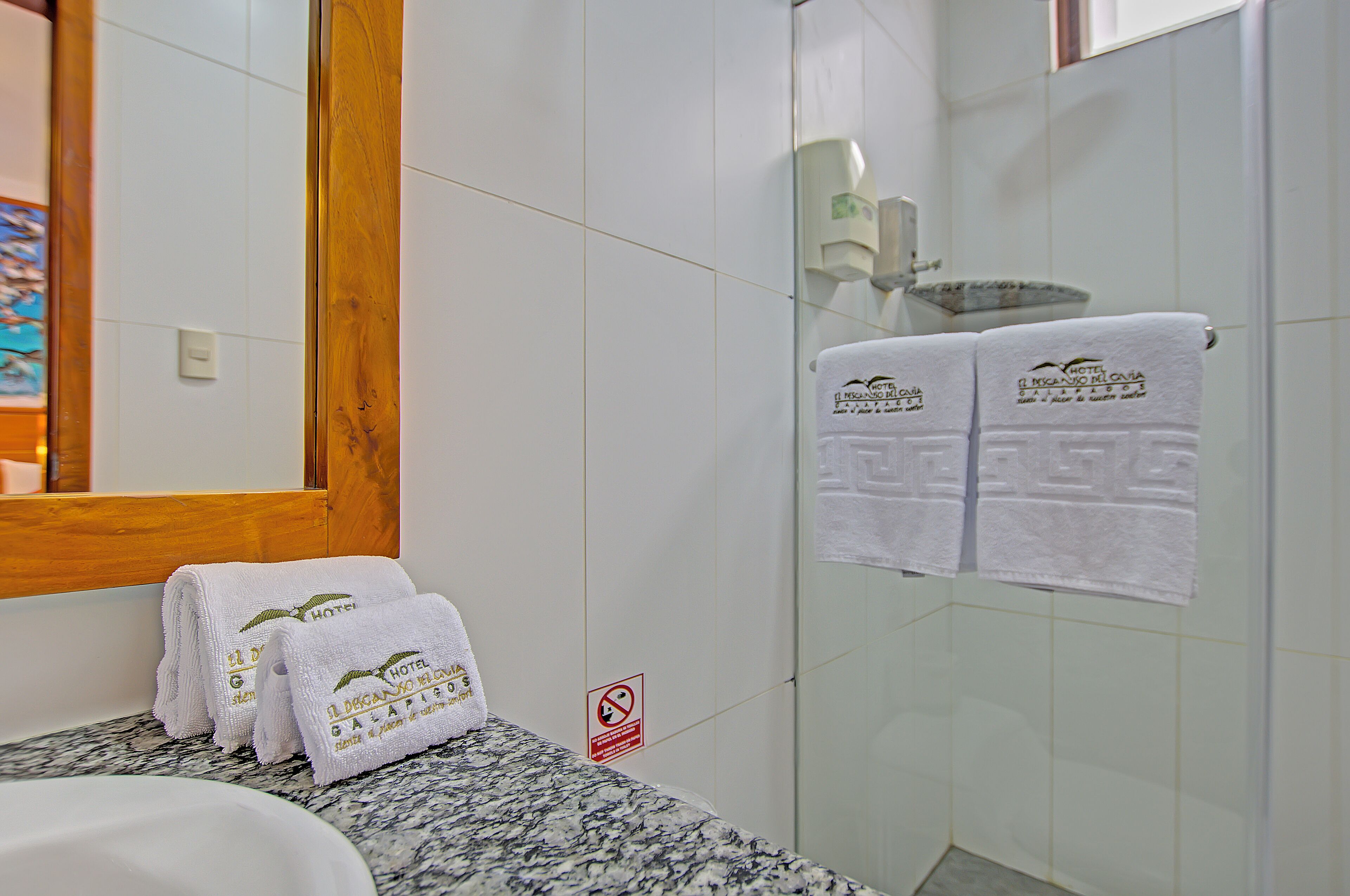 Uma casa de banho com lavatório de mármore, duas toalhas brancas com a marca do hotel, cabine de duche em vidro e espelho com moldura de madeira.